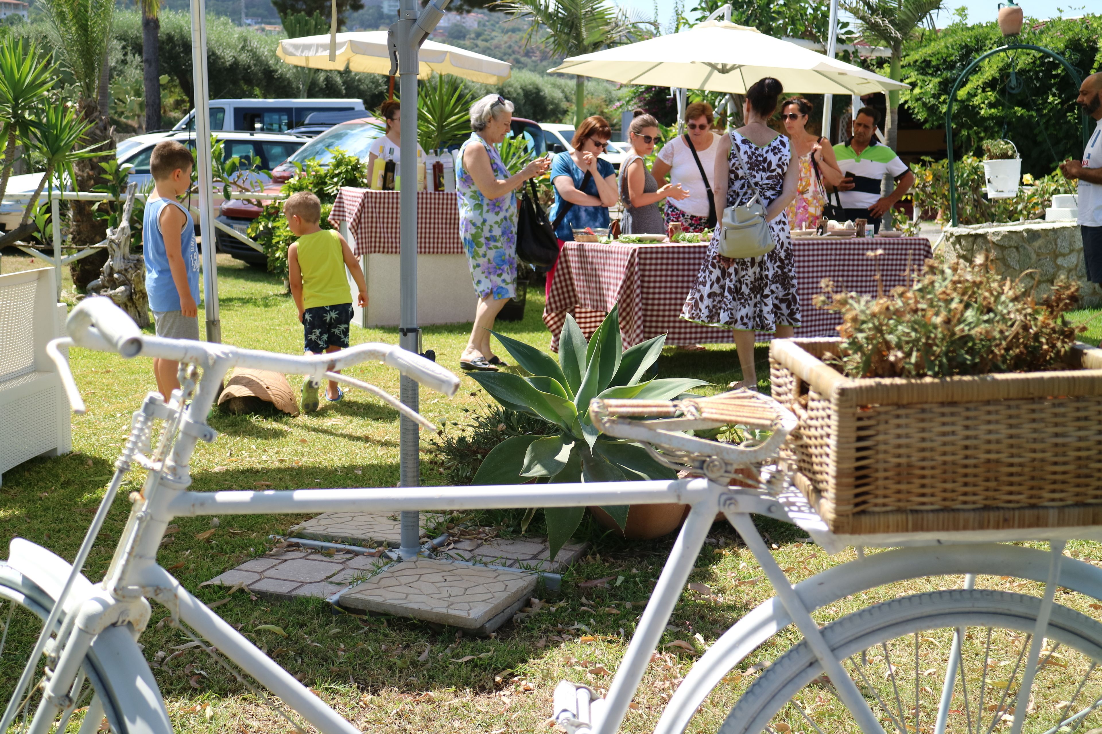 Uma bicicleta branca com cesto em primeiro plano, uma mesa com toalha xadrez e pessoas em pé em área externa com gramado.
