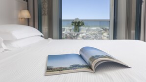 Quarto de hotel com uma cama, janela com vista para um prédio e um vaso de flores, além de uma revista sobre a cama.