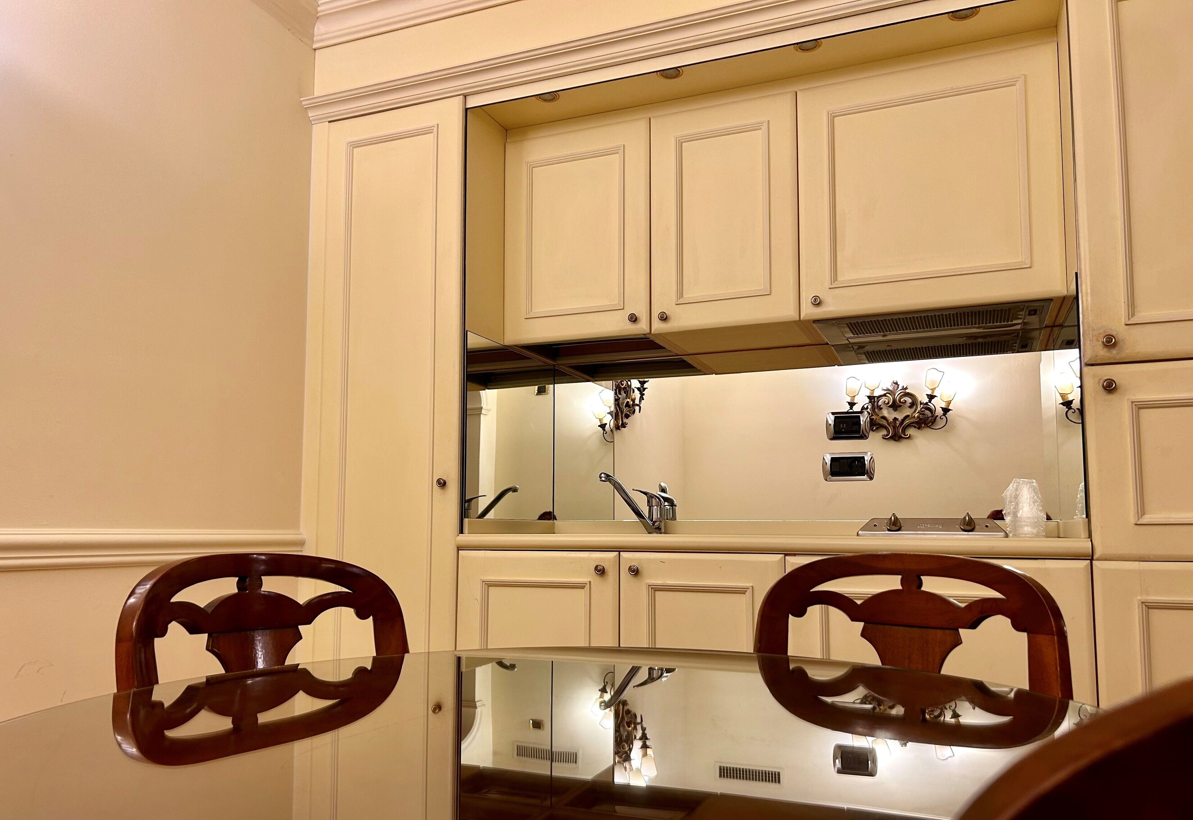 Uma área de refeições com mesa de vidro e cadeiras de madeira, uma cozinha cor creme com armários e um forno embutido.