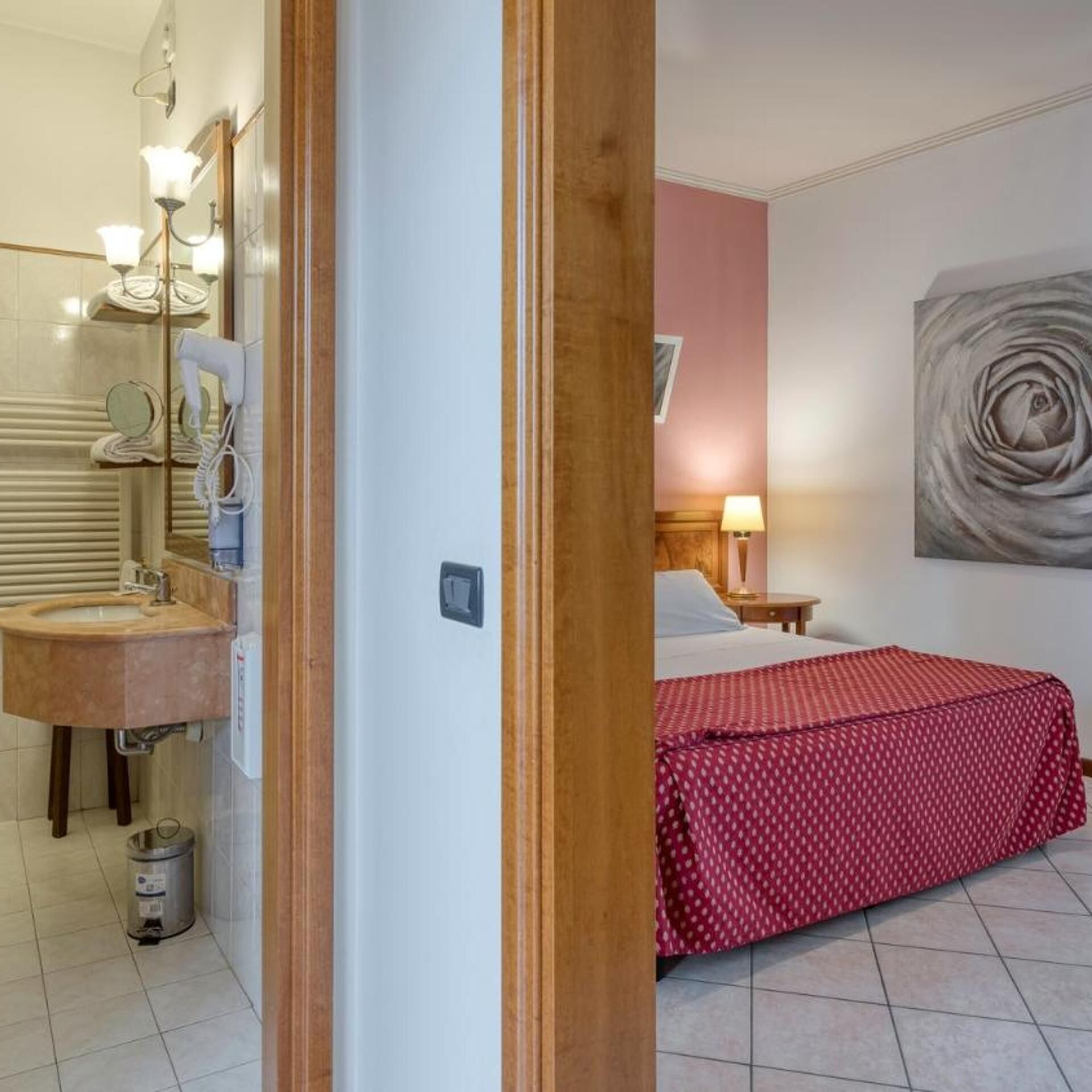 Quarto de hotel com banheiro, uma cama com colcha vermelha de bolinhas, um quadro na parede e um porta de madeira.