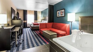 Quarto de hotel com cama, sofá vermelho, escrivaninha com cadeira, banheiro com banheira e uma grande janela com cortinas.