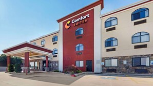 Um hotel Comfort Suites com fachada vermelha e bege, uma entrada coberta e céu claro.
