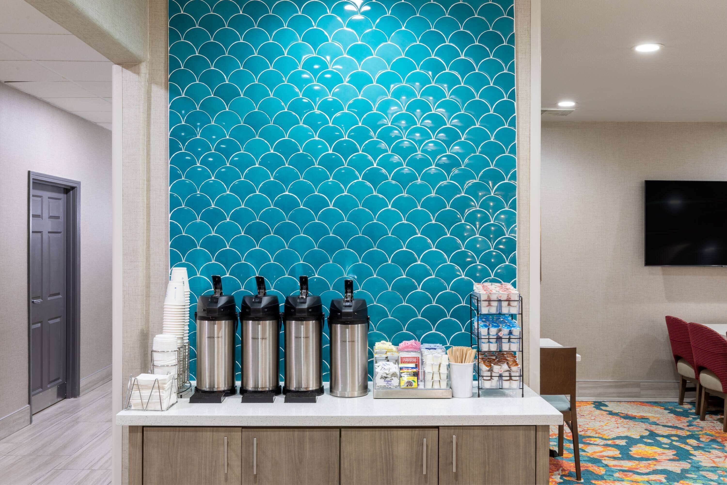 Uma área de recepção de hotel com parede em azulejos azuis, um balcão com máquinas de café e uma televisão fixada na parede.