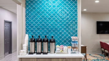 Uma área de recepção de hotel com parede em azulejos azuis, um balcão com máquinas de café e uma televisão fixada na parede.