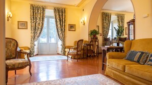 Uma sala de estar espaçosa com um sofá amarelo, uma mesa de centro de madeira e um tapete floral. Há duas poltronas e uma mesa de jantar com cadeiras. O quarto tem uma janela grande com cortinas, um espelho e luminárias na parede.