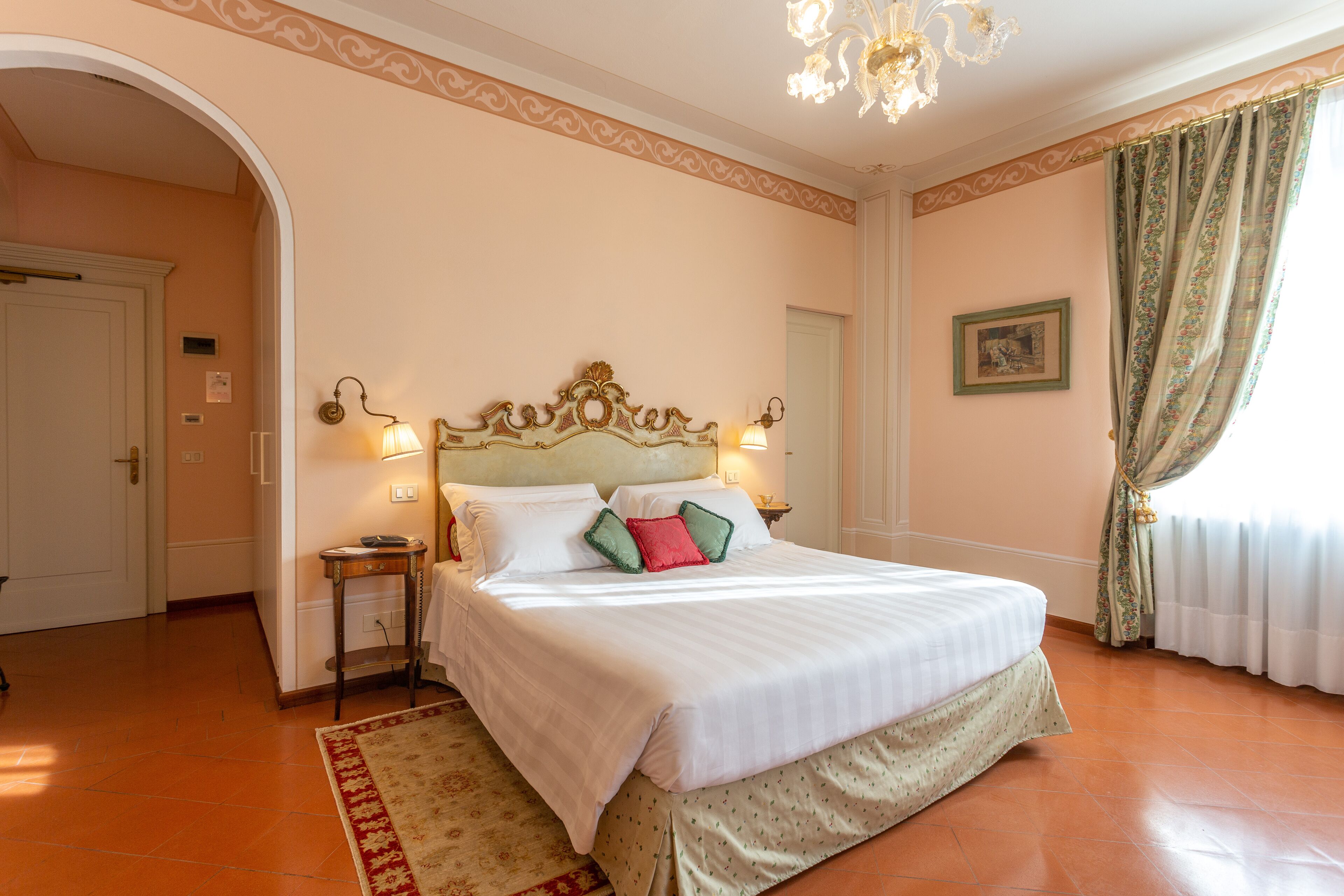 Quarto de hotel com uma cama grande, um candelabro, uma pequena mesa e uma porta que dá para outro quarto.