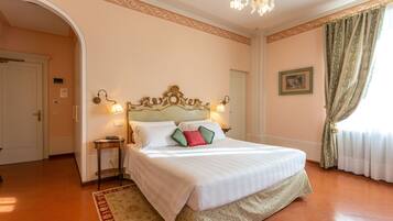 Quarto de hotel com uma cama grande, um candelabro, uma pequena mesa e uma porta que dá para outro quarto.