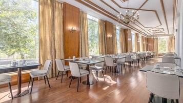 Uma sala de jantar ampla, com várias mesas e cadeiras, grandes janelas com cortinas e um lustre.