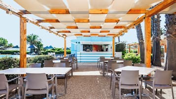 Um café ao ar livre com mesas e cadeiras, um bar ao fundo e palmeiras.