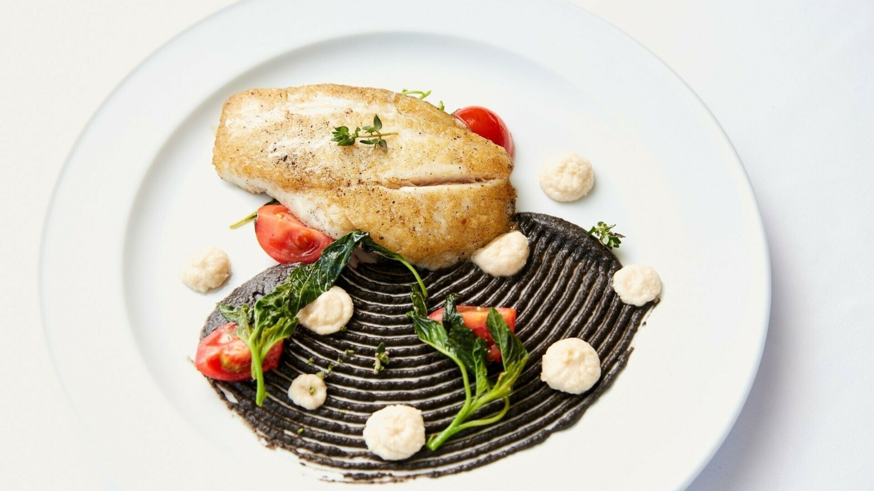 Um prato com um pedaço de peixe, tomates cereja e um molho escuro com padrão circular.
