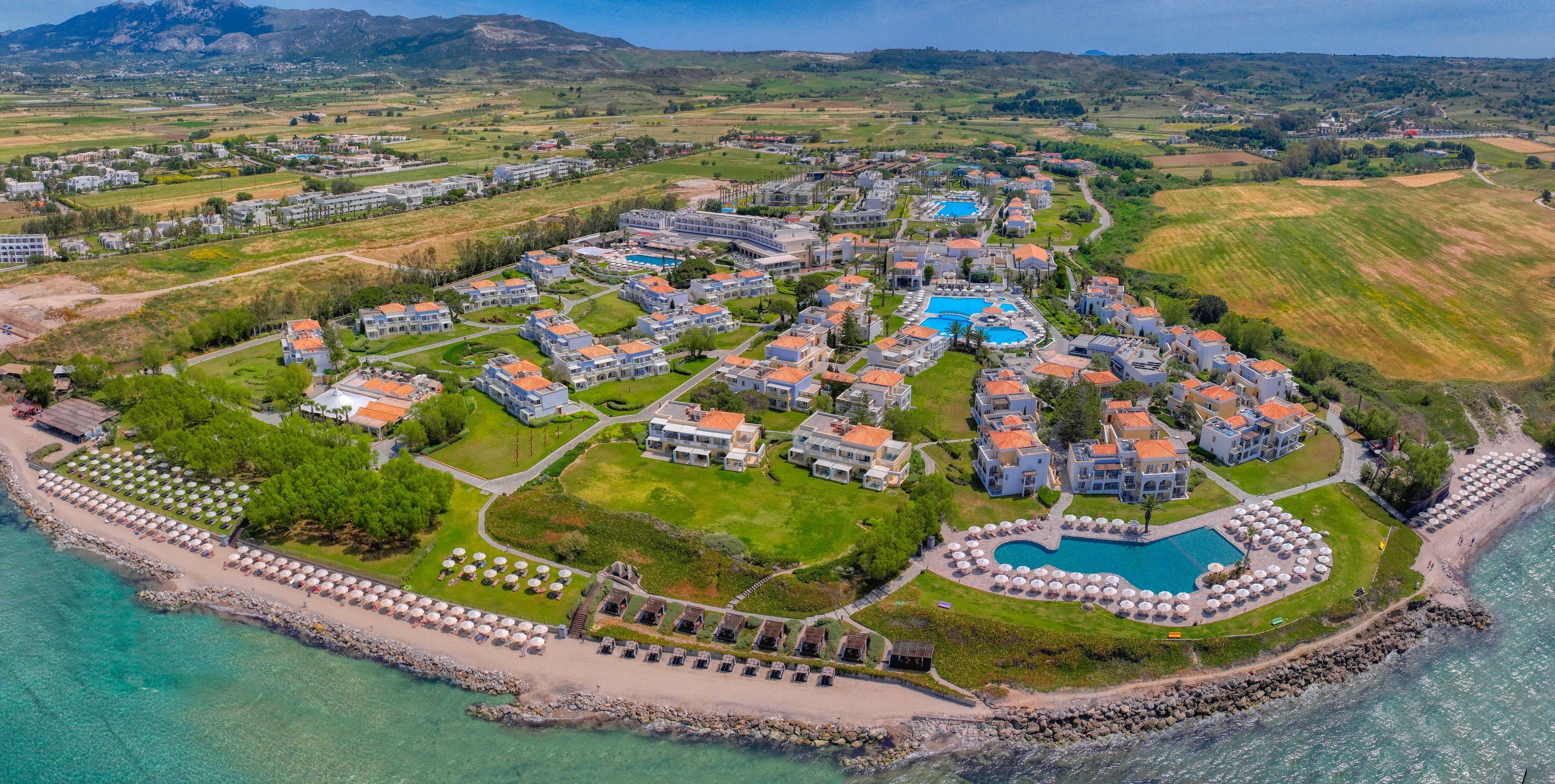 Vista aérea de um resort à beira-mar, com vários edifícios, uma piscina central e uma praia com guarda-sóis.