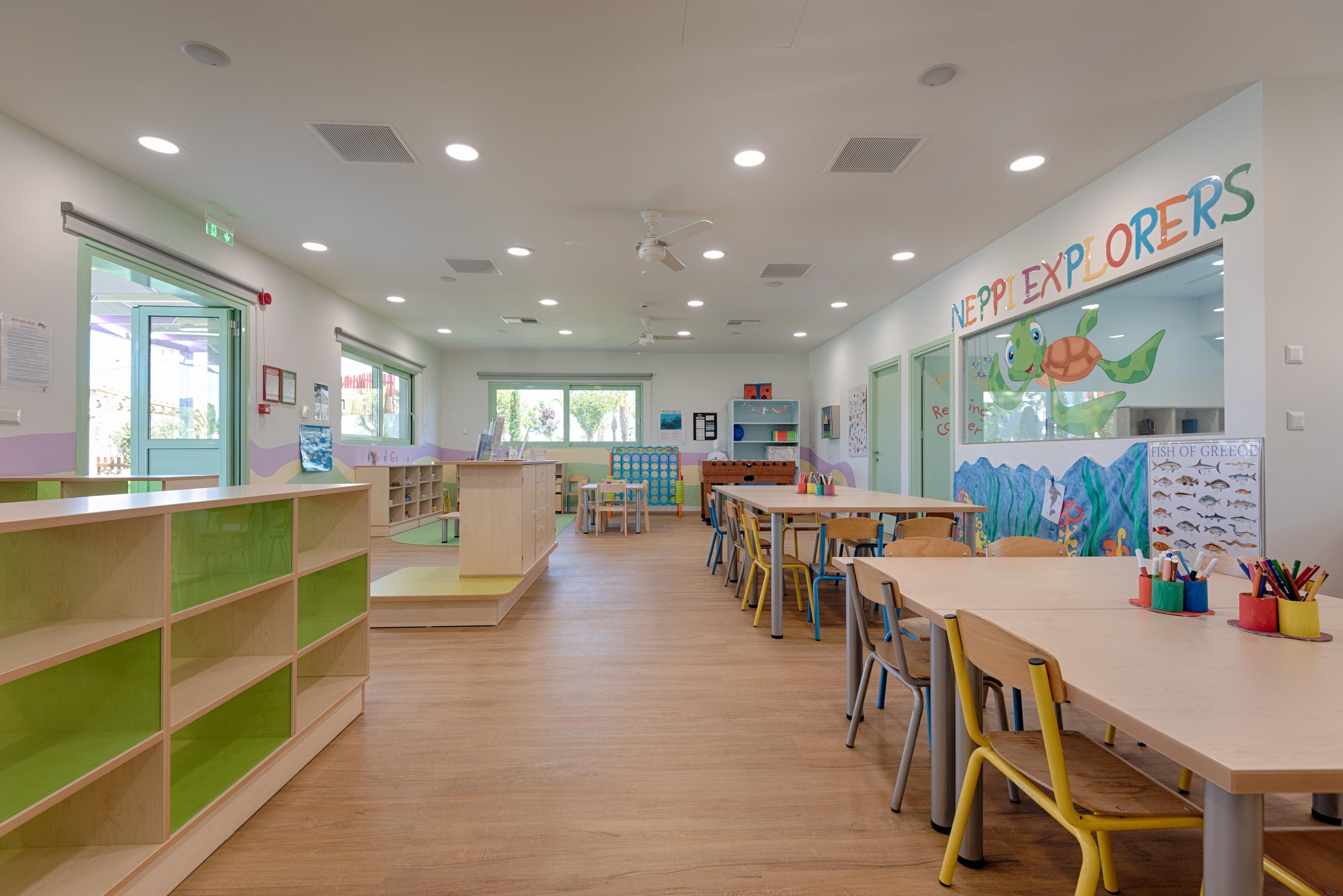 Uma sala de aula com mobiliário de madeira, um mural de tartaruga e cartazes educativos.