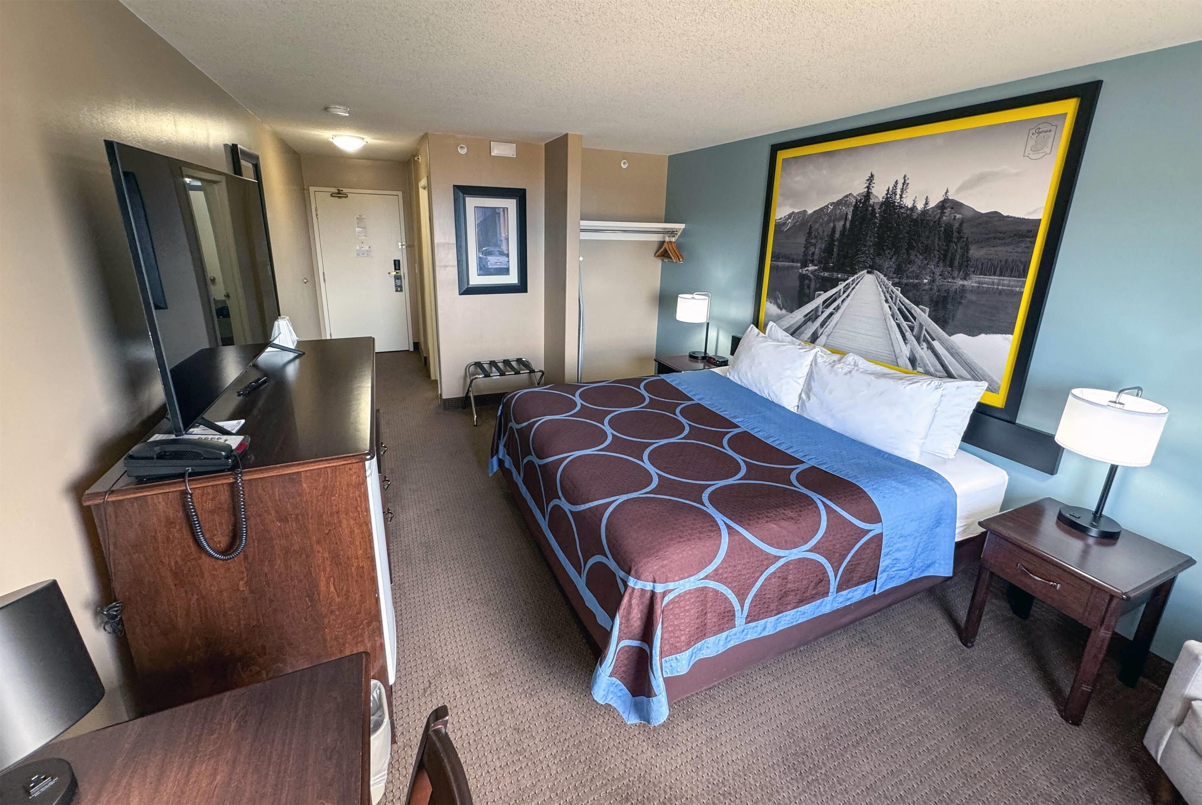 Quarto de hotel com cama, escrivaninha, penteadeira, espelho, quadro na parede e porta que dá para outro cômodo.