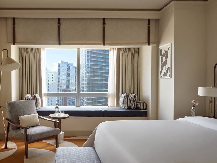 Quarto de hotel com uma cama grande, área de estar com uma poltrona e uma mesinha, janela com cortinas e vista para a cidade.