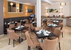 Uma sala de jantar de hotel moderna com várias mesas e cadeiras, uma televisão fixada na parede e uma área de cozinha visível ao fundo.