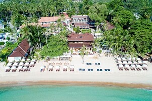 Um hotel à beira-mar com vários edifícios, palmeiras e uma praia de areia com espreguiçadeiras.