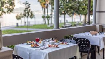 Área de refeições ao ar livre com mesas preparadas para o café da manhã, com vista para um corpo d'água e vegetação.