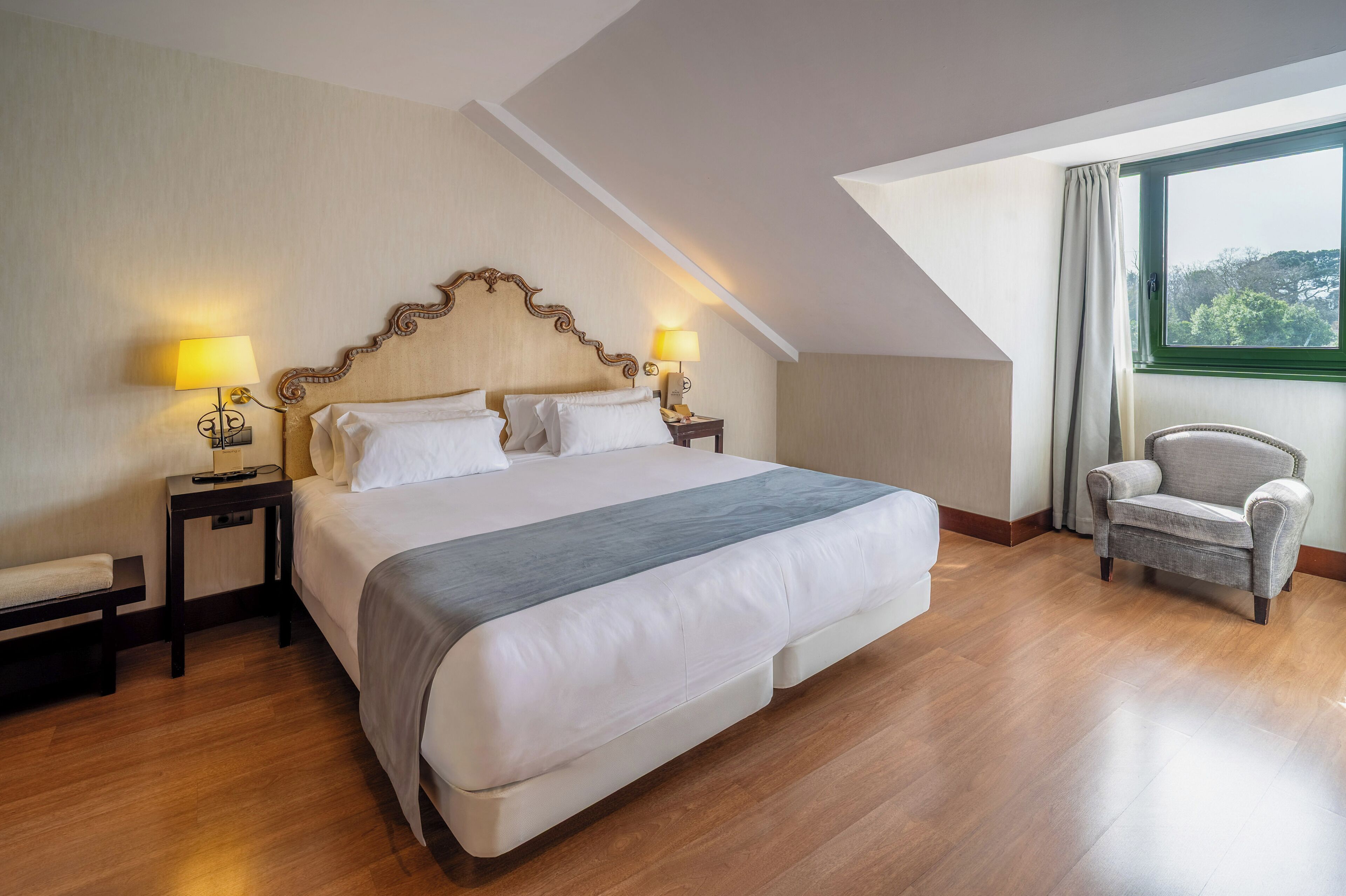 Quarto com cama grande, duas mesas de cabeceira com luminárias, um banquinho, uma cadeira e uma janela com cortinas.