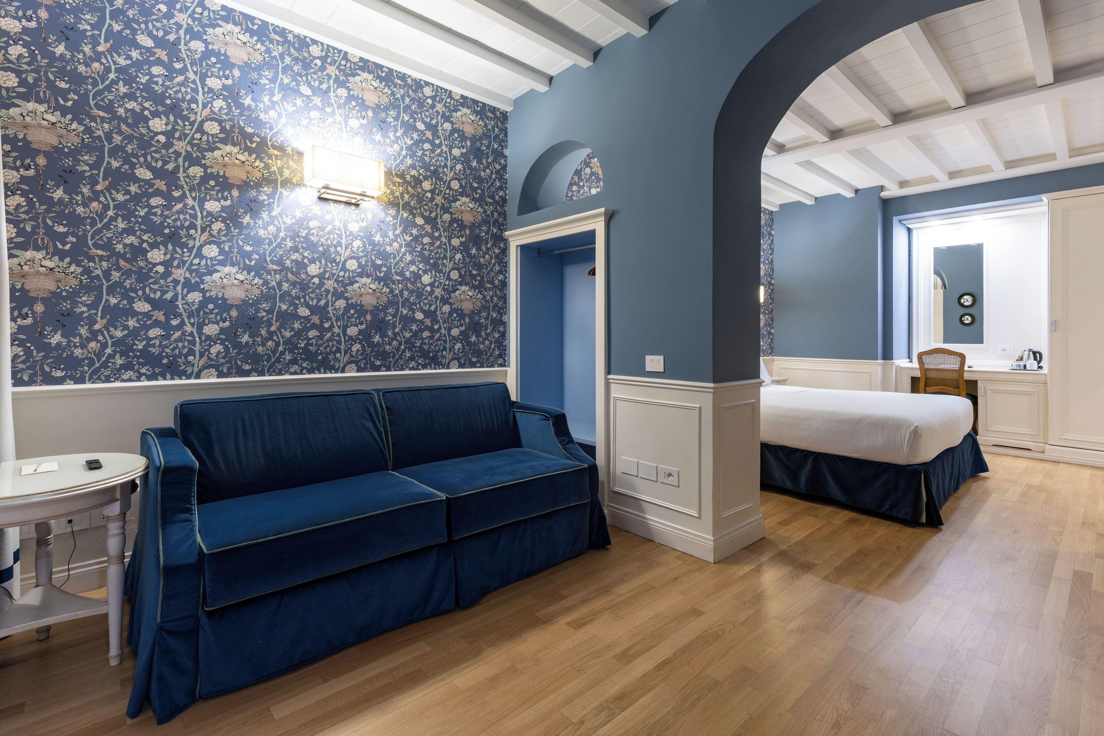 Quarto com papel de parede floral azul, um sofá azul, piso de madeira e cama com roupa de cama branca.