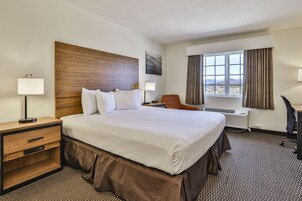 Quarto de hotel com uma cama grande, uma escrivaninha, uma cadeira, uma mesa de cabeceira, um abajur e uma janela com cortinas.