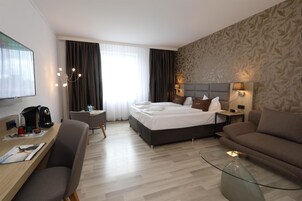 Quarto de hotel com cama, escrivaninha com cafeteira, sofá, televisão e janela com cortinas.