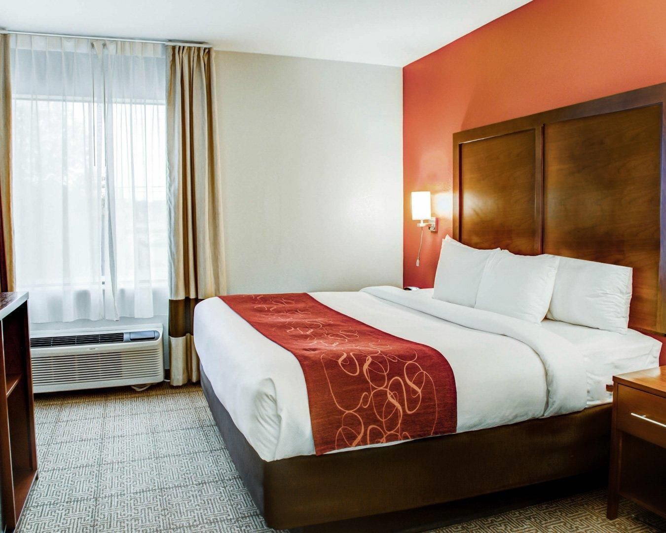 Quarto de hotel com cama grande, mesa de cabeceira, abajur, janela com cortinas e ar condicionado.