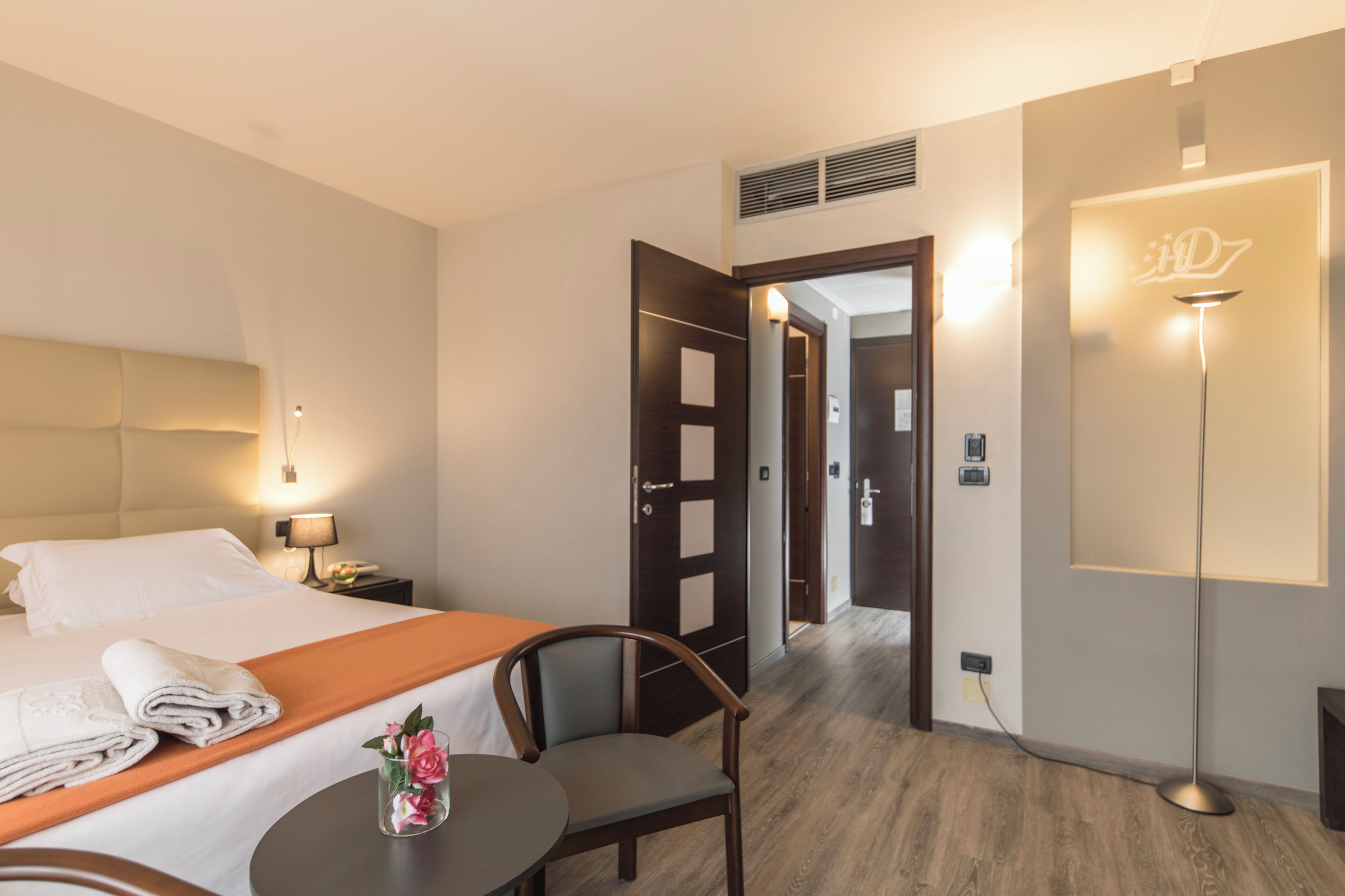 Quarto de hotel com cama, mesa de cabeceira, cadeira e uma pequena mesa com um vaso de flores.