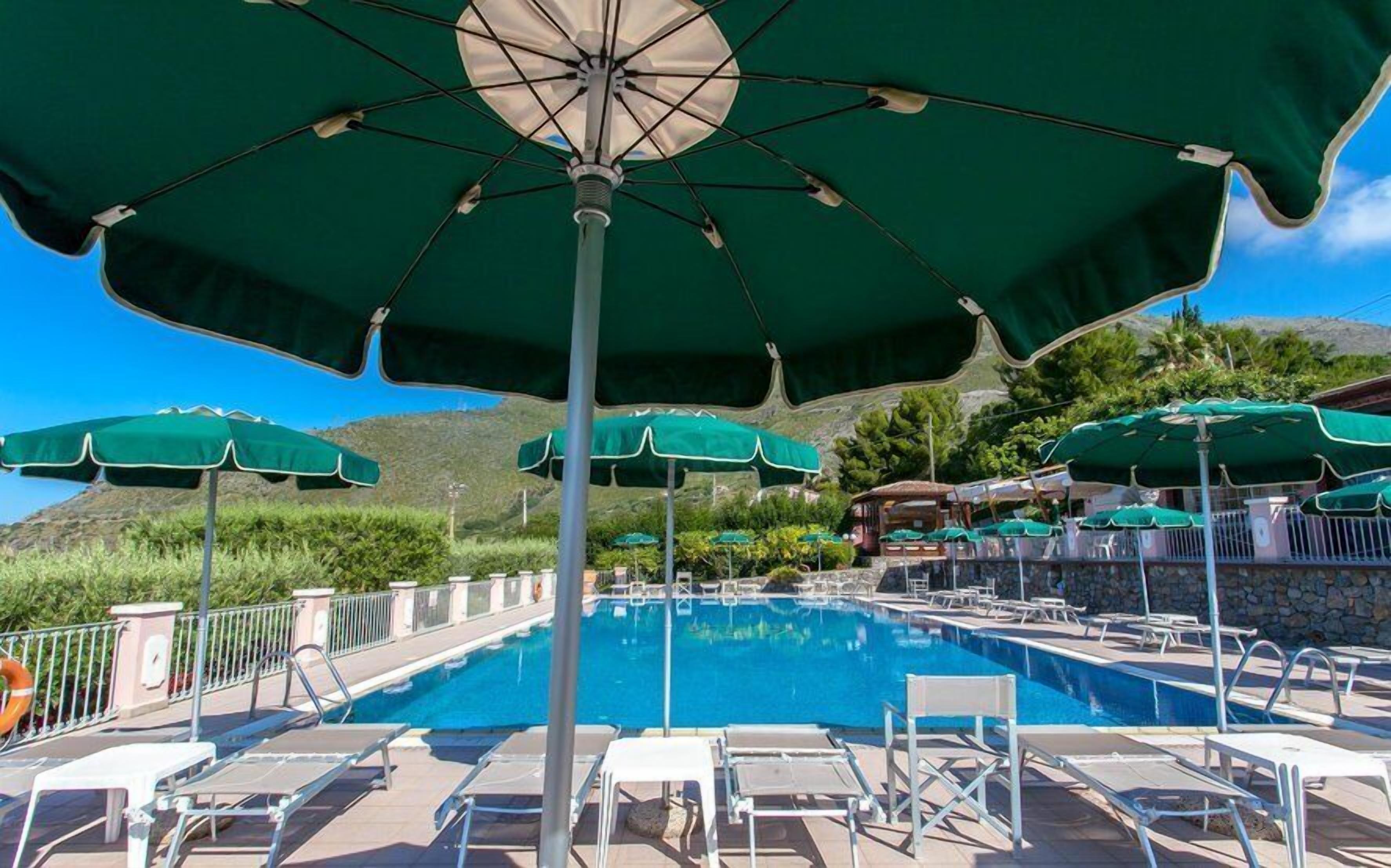Uma área de piscina com vários guarda-sóis verdes, cadeiras de descanso e uma vista desimpedida da paisagem ao redor.