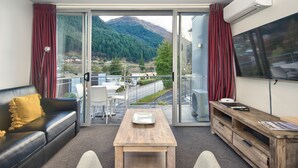 Uma sala de estar moderna com sofá, mesa de madeira e televisão. A sala possui grandes portas de vidro que dão para uma varanda com vista para as montanhas.