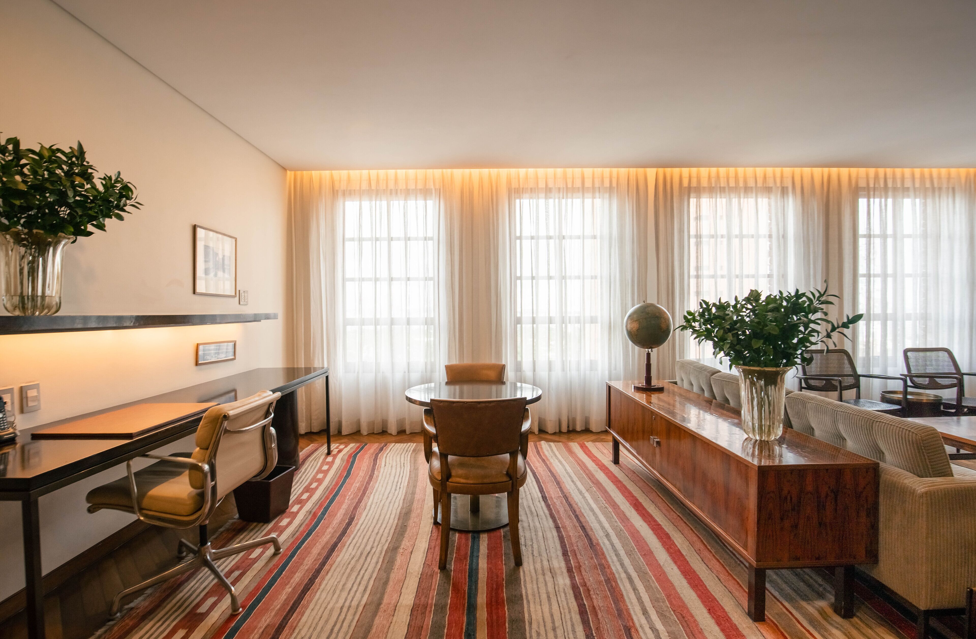 Quarto de hotel com mesa de jantar, cadeiras, sofá, escrivaninha e uma janela grande com cortinas.