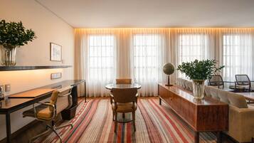 Quarto de hotel com mesa de jantar, cadeiras, sofá, escrivaninha e uma janela grande com cortinas.