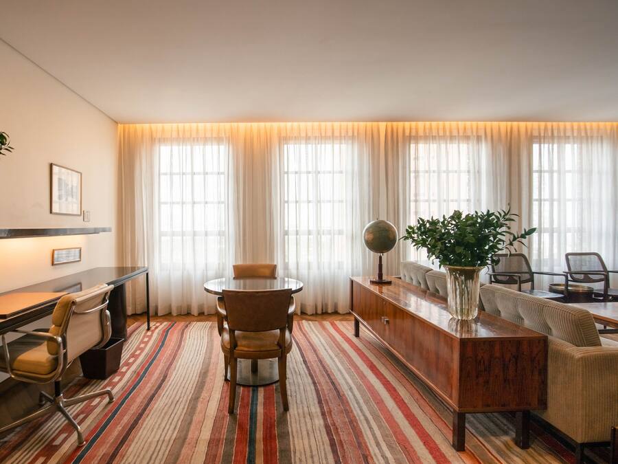 Quarto de hotel com mesa de jantar, cadeiras, sofá, escrivaninha e uma janela grande com cortinas.