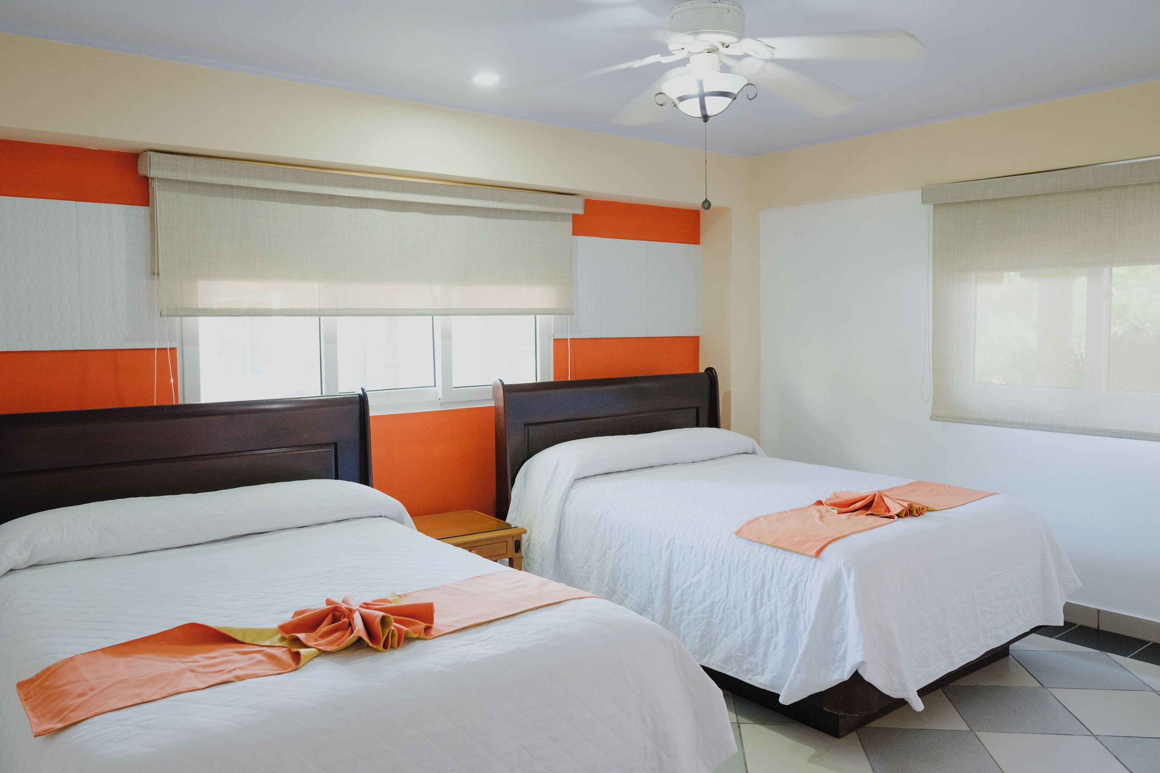 Quarto de hotel com duas camas, ventoinha no teto e janela com cortinas.