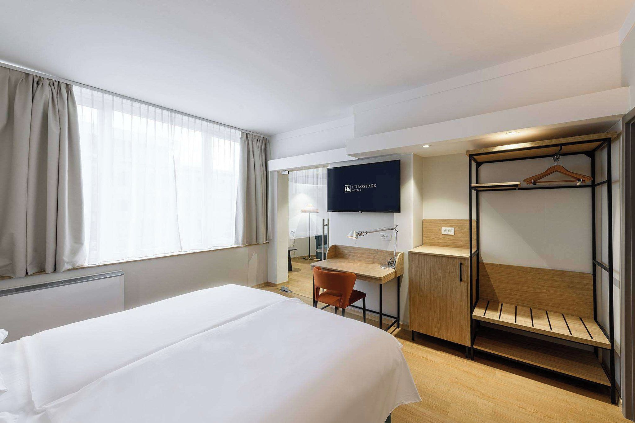Quarto de hotel com cama, escrivaninha, cadeira, televisão e closet espaçoso.
