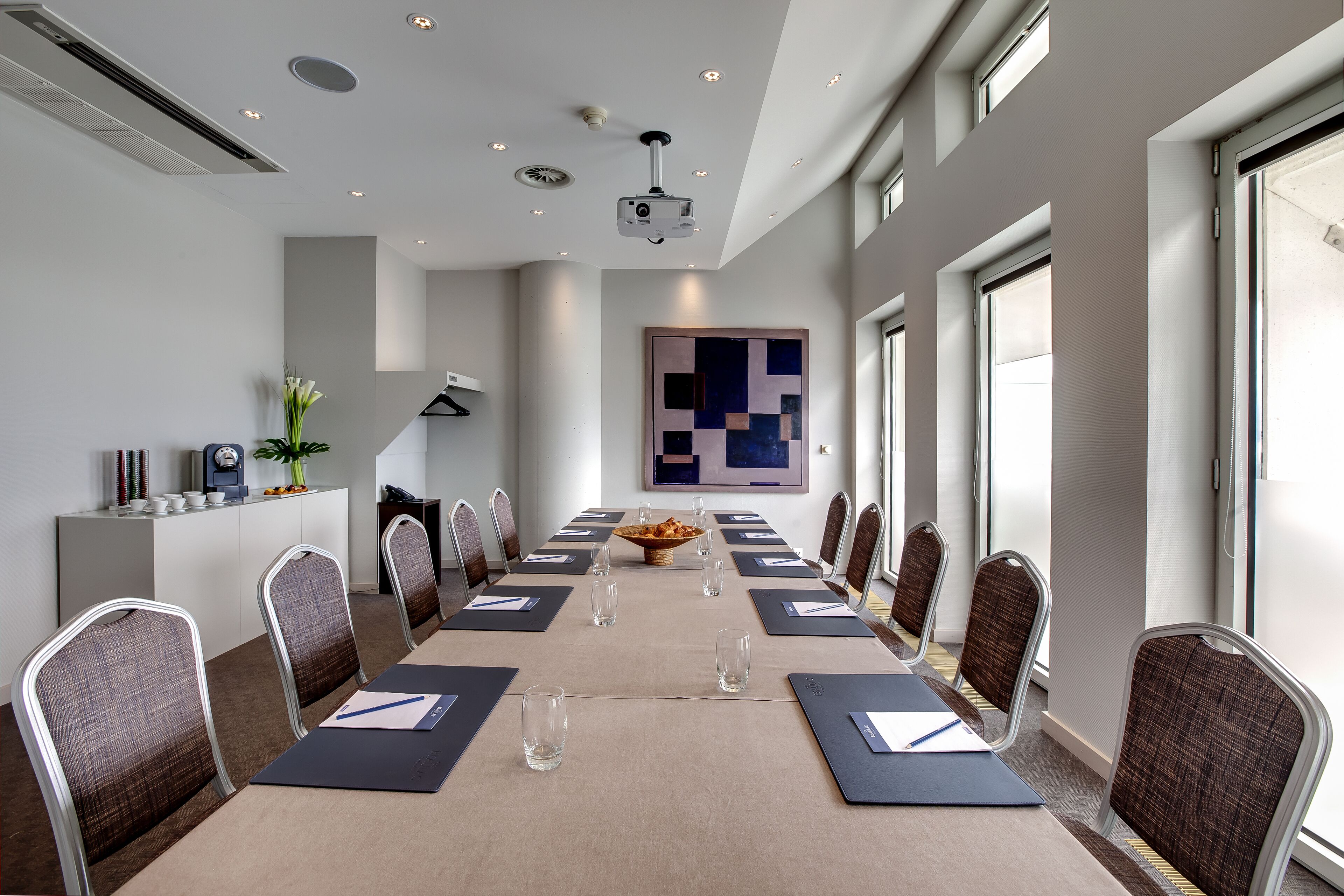 Uma sala de conferência com uma mesa comprida, cadeiras, um projetor e um grande quadro abstrato na parede.