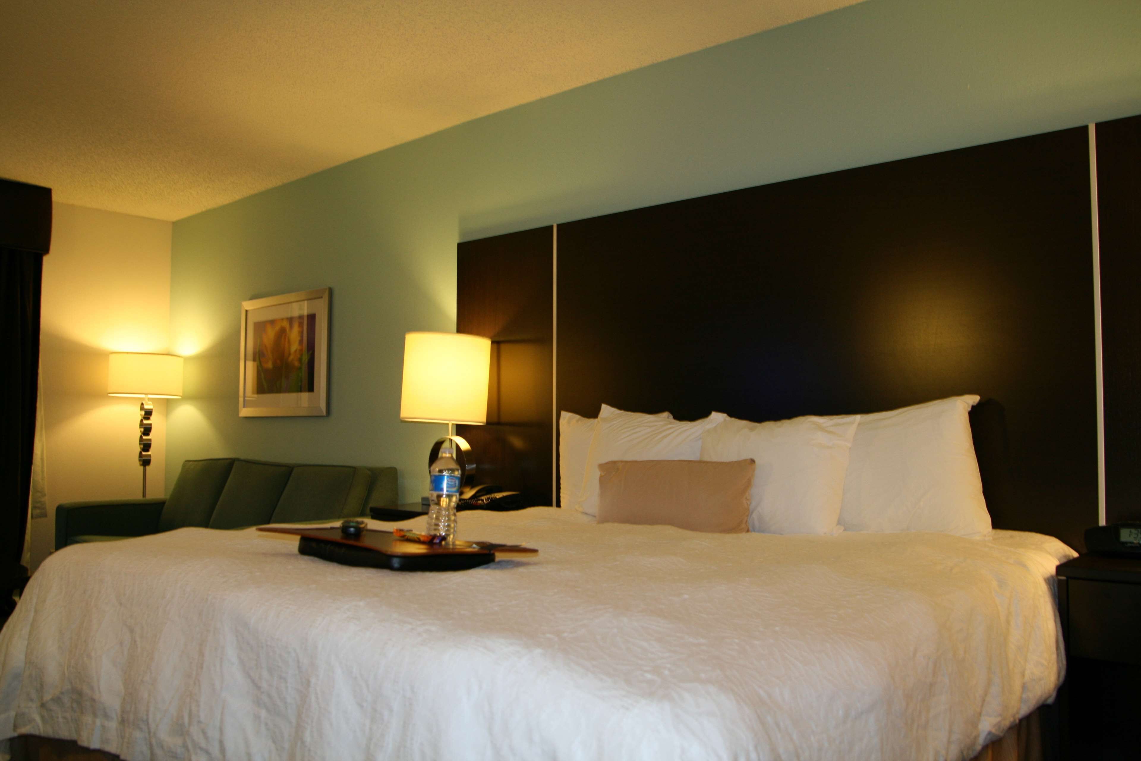 Quarto de hotel com uma cama grande, duas luminárias de cabeceira, uma mesa de cabeceira e uma poltrona verde.