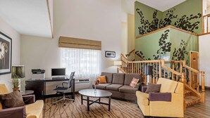 Uma sala ampla com um sofá moderno, uma mesa de centro redonda e uma escrivaninha com um computador. Uma escada com corrimãos de madeira leva a um andar superior.