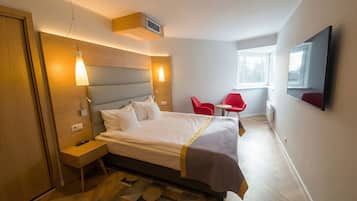 Quarto de hotel com cama, mesas de cabeceira, uma cadeira vermelha, uma pequena mesa, televisão e janela.