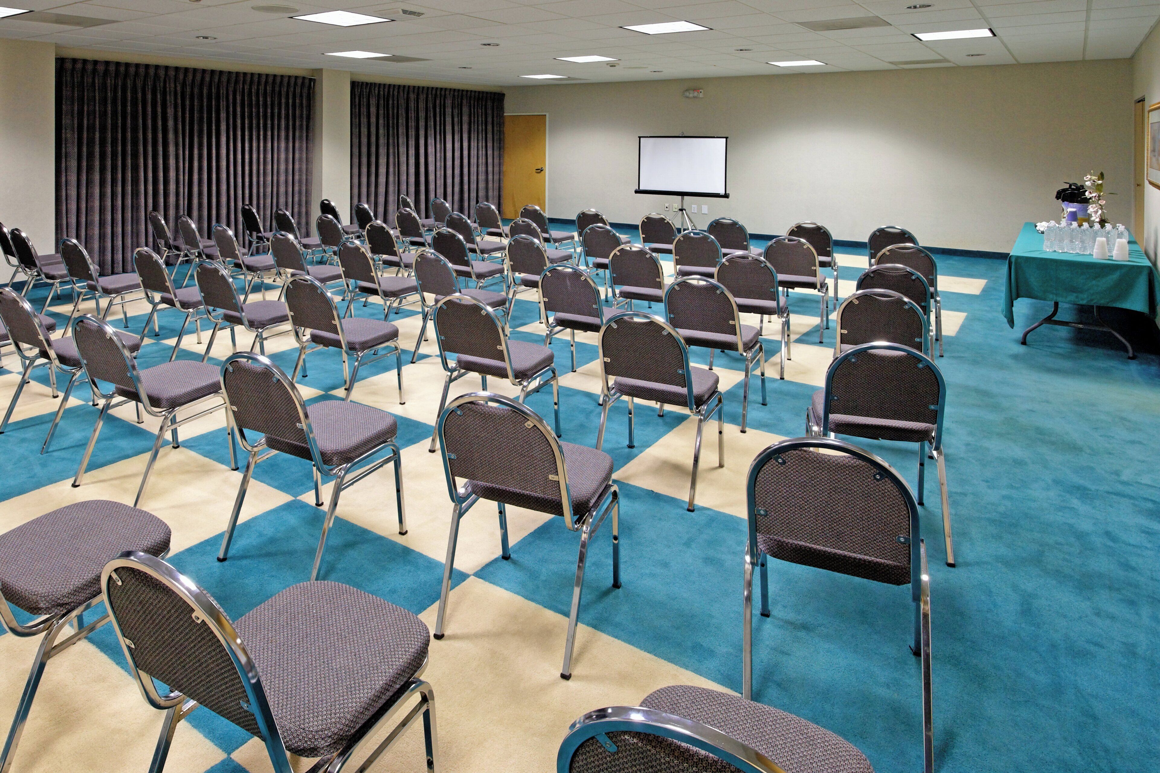 Uma sala de conferências com fileiras de cadeiras, uma tela de projetor e uma mesa com garrafas de água.