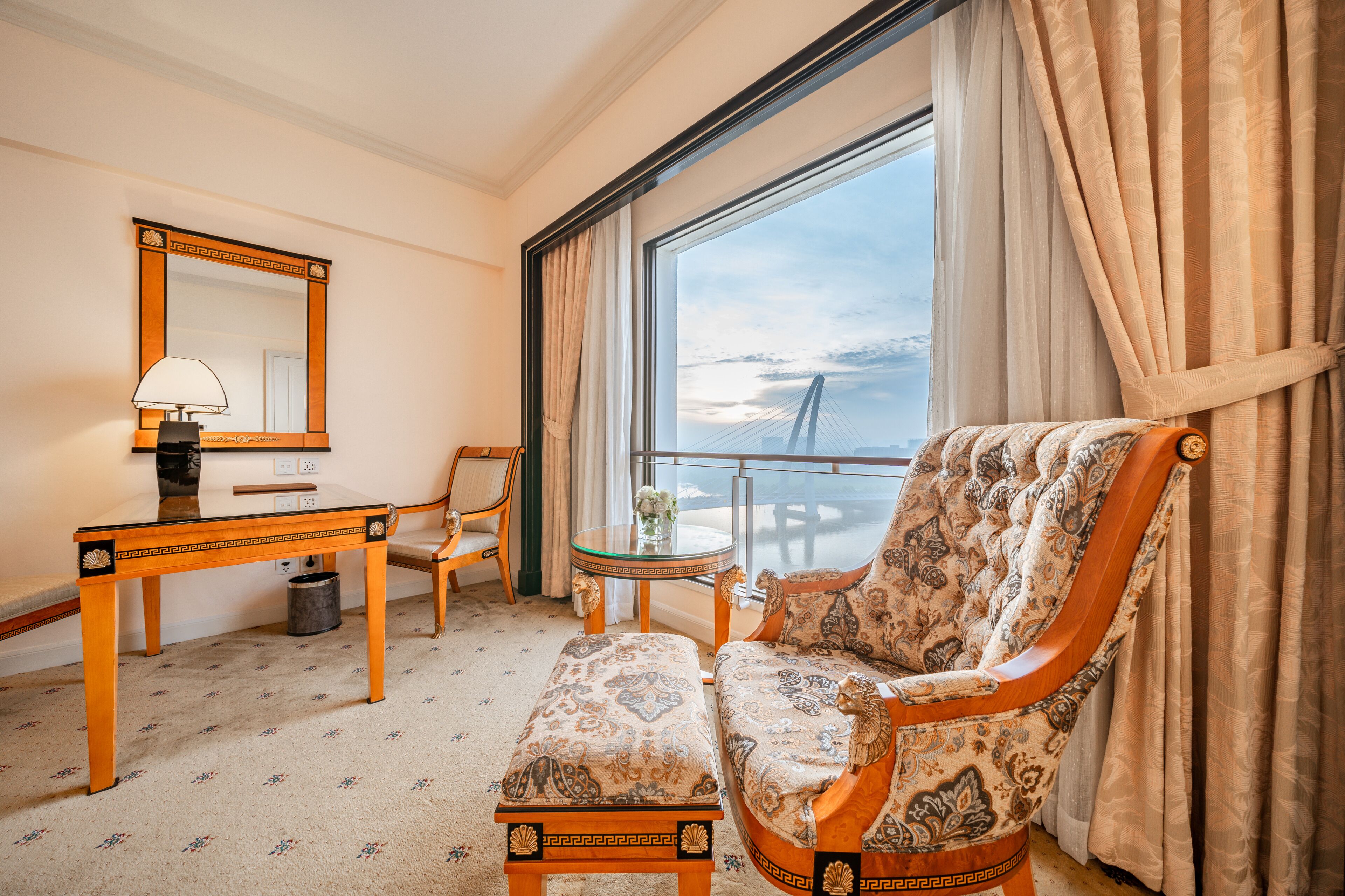 Quarto de hotel com uma grande janela, um sofá, uma cadeira, uma mesa pequena e um abajur.
