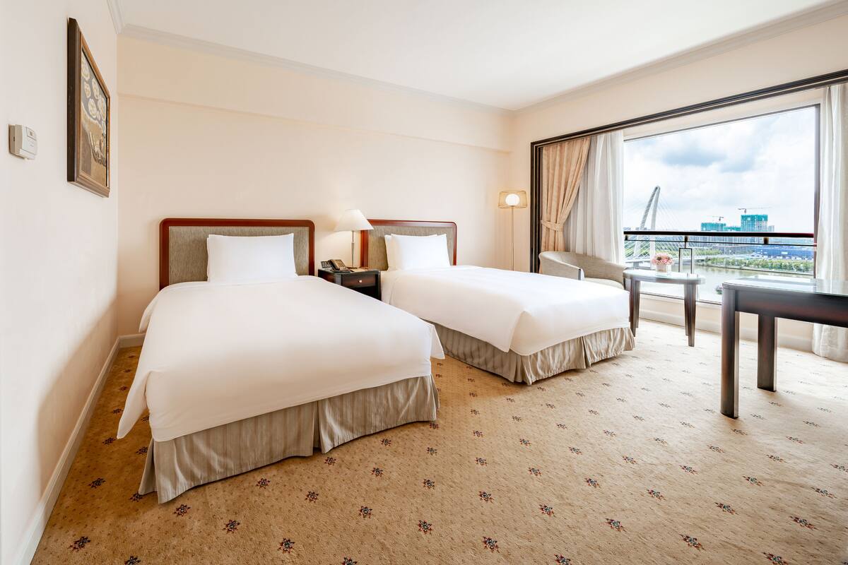 Quarto de hotel com duas camas, uma mesa, uma cadeira e uma janela grande com vista para uma ponte e paisagem urbana.