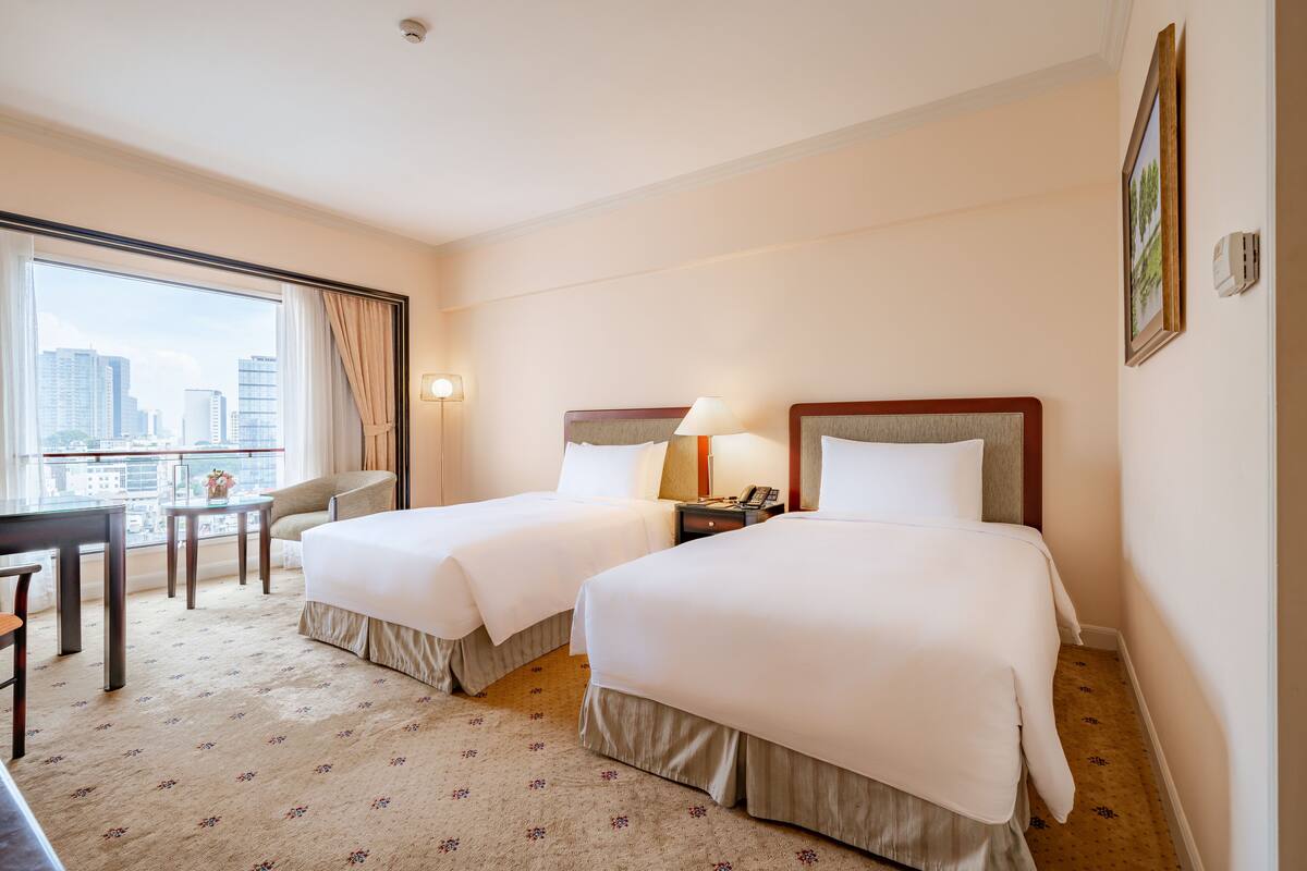 Quarto de hotel com duas camas, uma escrivaninha, uma cadeira, uma luminária e uma janela grande com vista para a cidade.