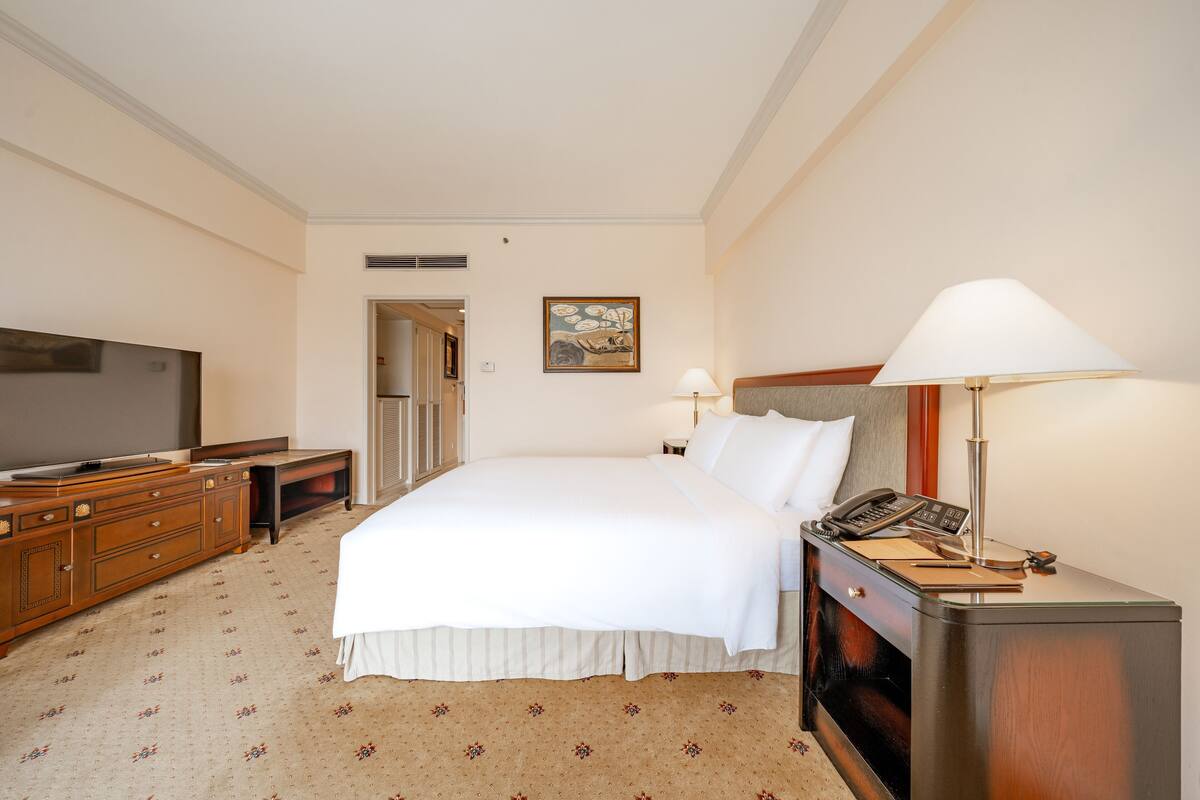 Quarto de hotel com cama, mesa de cabeceira, luminária, escrivaninha e televisão.