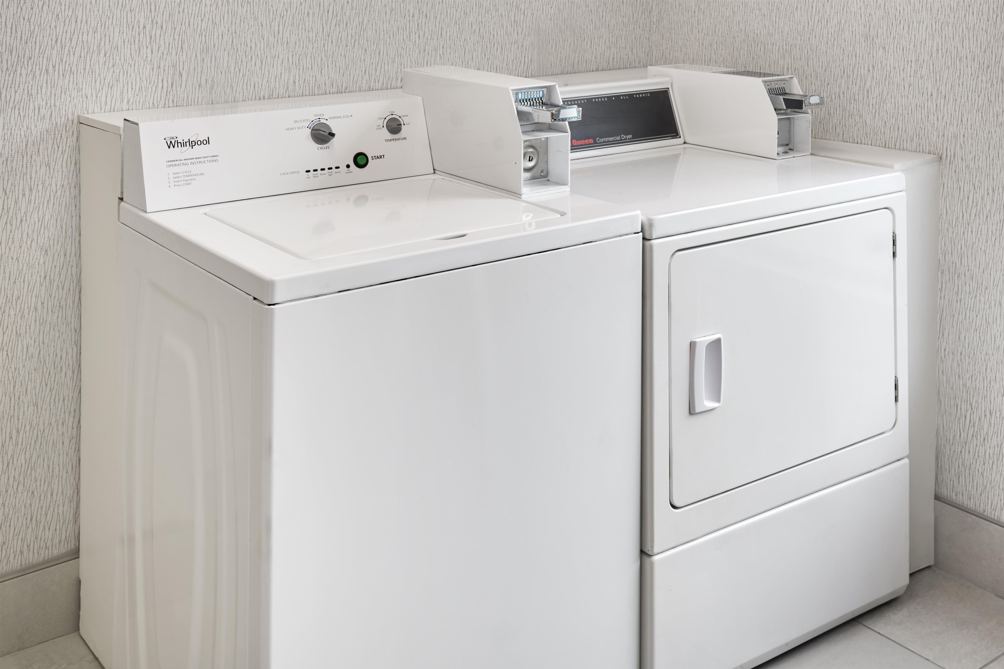 Máquina de lavar Whirlpool de abertura frontal, com painel de controle superior, porta com puxador e piso de azulejos brancos.