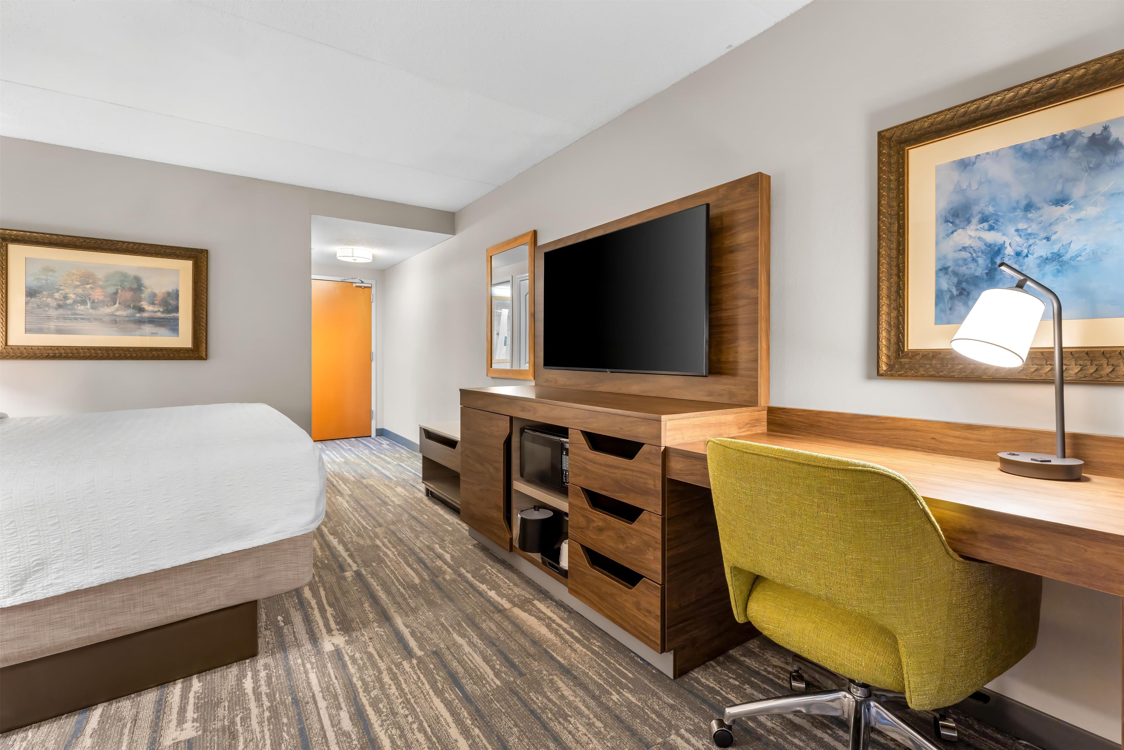 Quarto de hotel com cama, escrivaninha, cadeira, televisão e obras de arte nas paredes.