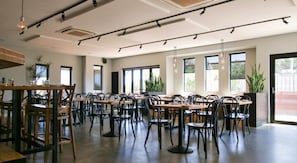 Uma ampla sala de jantar com várias mesas e cadeiras, grandes janelas e luminárias no teto.