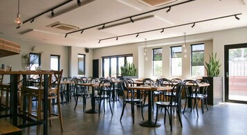 Uma ampla sala de jantar com várias mesas e cadeiras, grandes janelas e luminárias no teto.