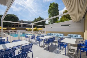 Uma área à beira da piscina com cadeiras e mesas azuis, uma piscina e um espaço de estar coberto com guarda-sóis amarelos.