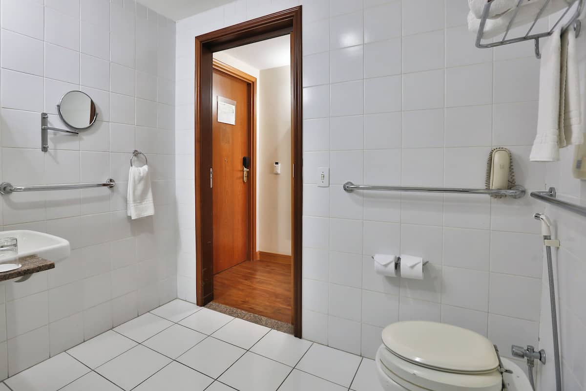 Um banheiro com azulejos brancos, uma porta de madeira, vaso sanitário, pia, espelho, porta-toalhas e área de chuveiro.