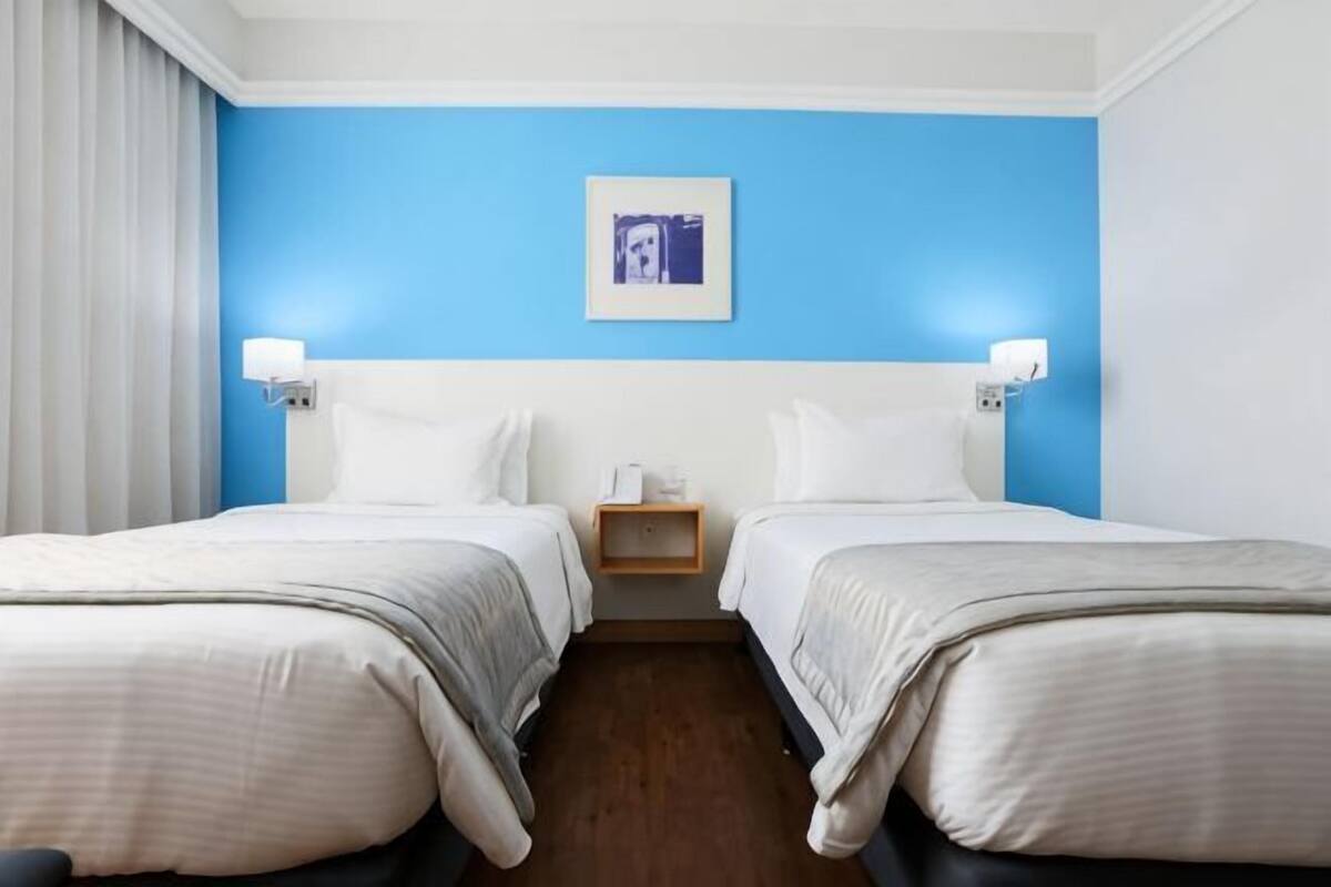 Quarto de hotel com duas camas, parede azul decorativa, mesa de cabeceira com telefone e quadro na parede.